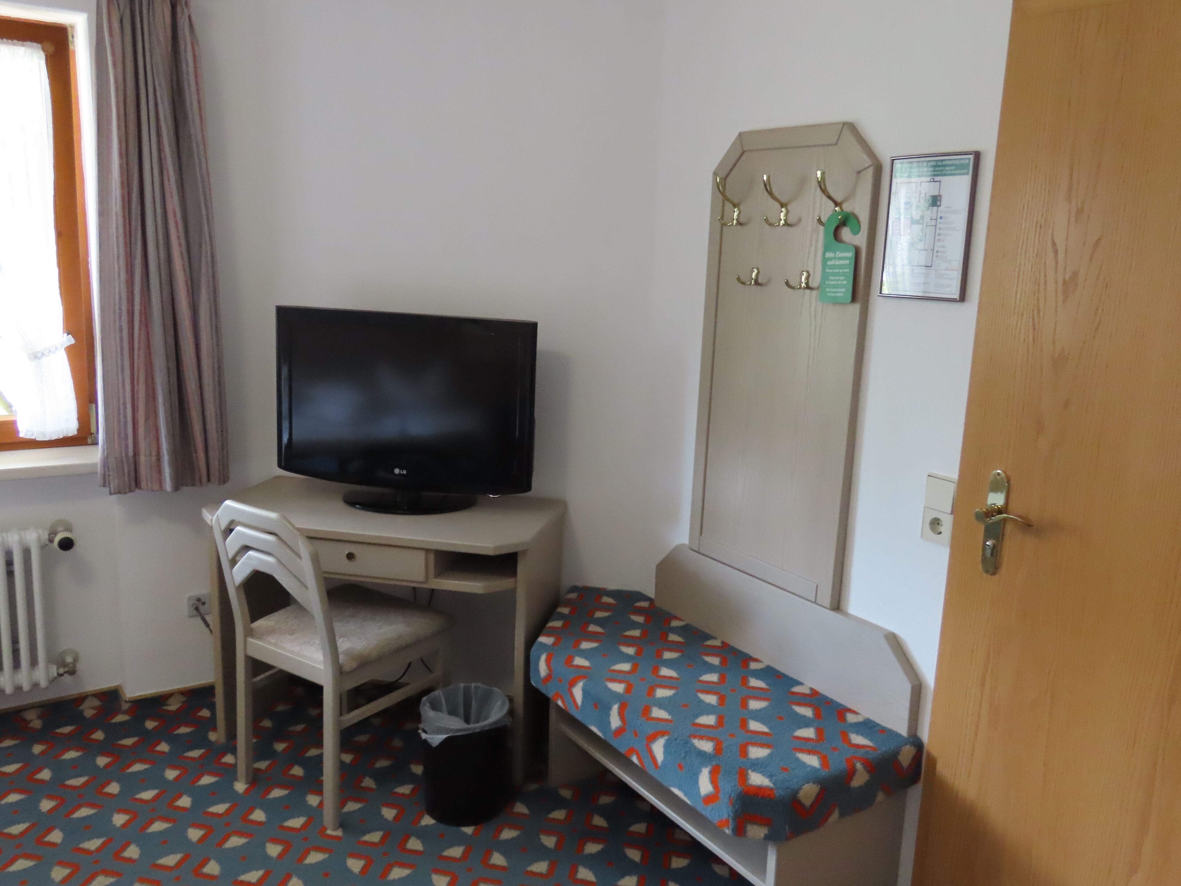 Quarto pequeno com uma cama, escrivaninha com televisão, cadeira, radiador e porta.