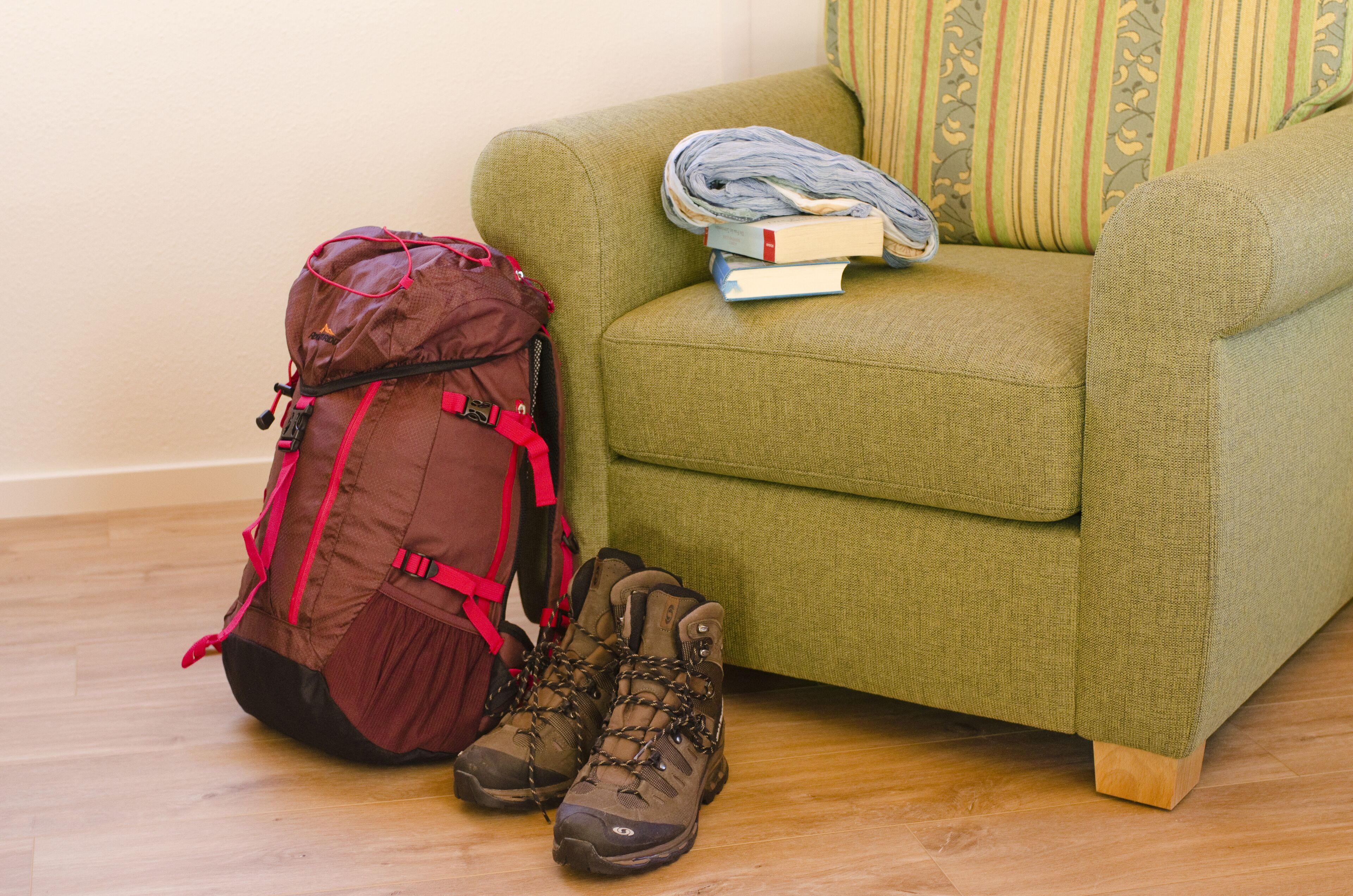 Um sofá verde com uma manta enrolada e um livro, uma mochila marrom e um par de botas de caminhada.
