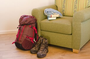 Um sofá verde com uma manta enrolada e um livro, uma mochila marrom e um par de botas de caminhada.