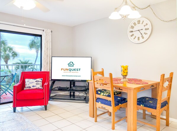 Um quarto com televisão exibindo 'FUNQUEST Vacation Rentals', uma mesa de jantar de madeira com cadeiras, uma poltrona vermelha e vista para palmeiras pela janela.