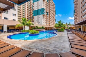 Uma área de piscina de hotel com espreguiçadeiras, piscina e edifícios altos ao fundo.