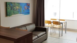 Uma sala de estar moderna com um sofá de couro, uma mesa de centro de madeira e um quadro representando uma paisagem tropical. Há uma área de jantar com mesa e cadeiras.
