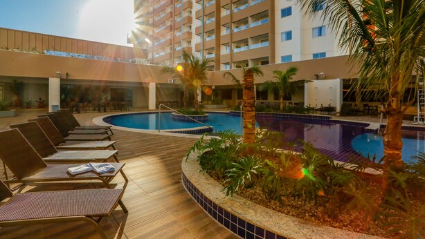 Uma área de piscina com espreguiçadeiras, palmeiras e um prédio alto ao fundo.