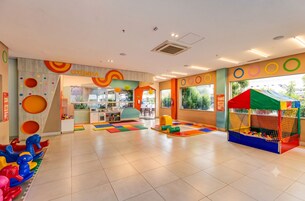 Espaço de brincadeiras interno, bem iluminado, com uma tenda de jogos, tapetes coloridos e uma área de cozinha com micro-ondas.