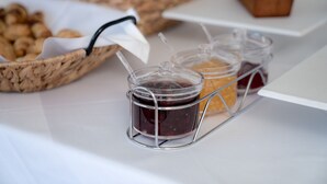 Um buffet de café da manhã com uma cesta de vime de doces, dois recipientes de vidro para acompanhamentos e uma baguete em um prato branco.