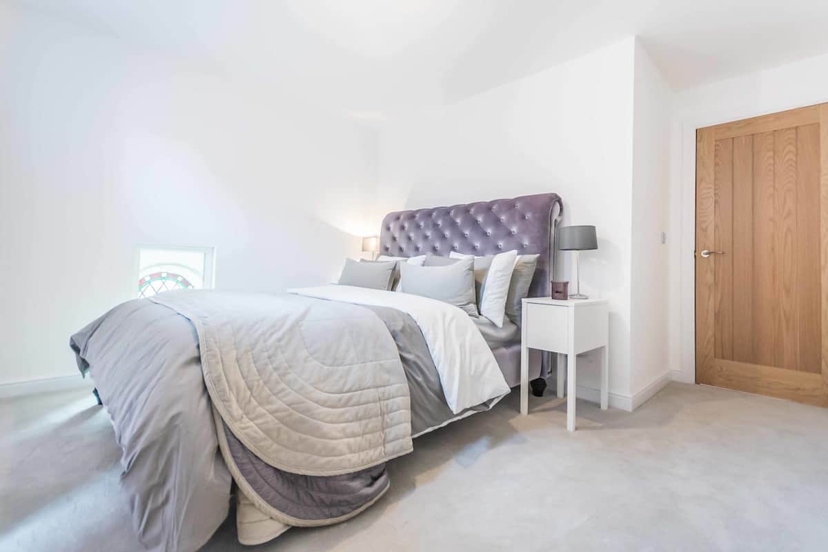 Quarto com cama, mesas de cabeceira, abajur e porta de madeira.