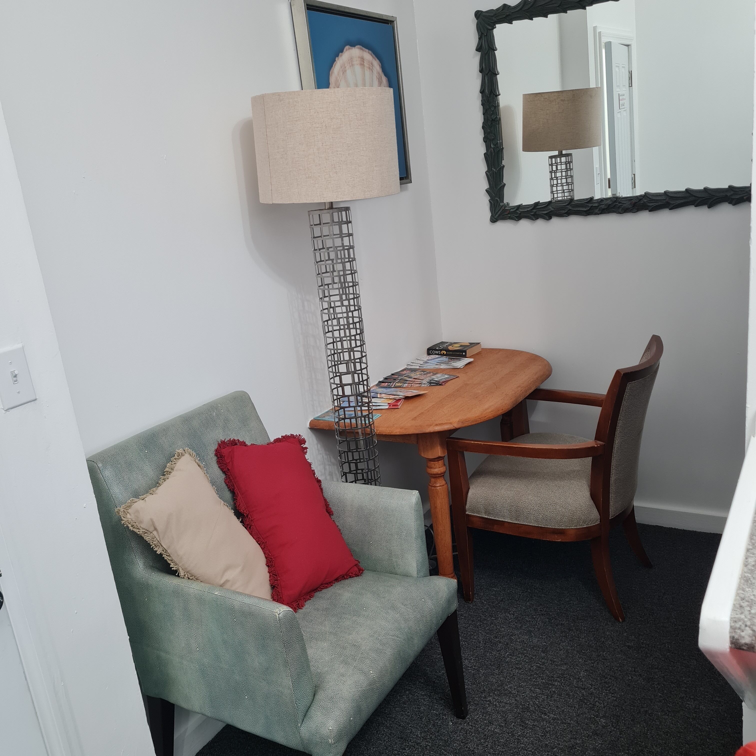 Um quarto pequeno com um sofá verde, uma mesa de jantar de madeira e uma cadeira. Há um abajur, um espelho e um quadro na parede.
