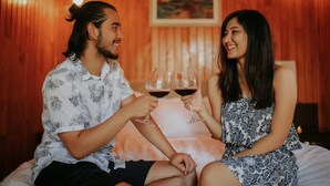 Um casal sentado em uma cama com cabeceira de madeira, segurando taças de vinho.