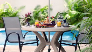 Mesa redonda com disposição para café da manhã à beira da piscina, incluindo cestas de pães, frutas e um bule de chá.