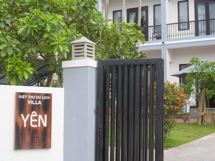 Uma villa com entrada protegida por portão, placa de identificação e um edifício ao fundo.