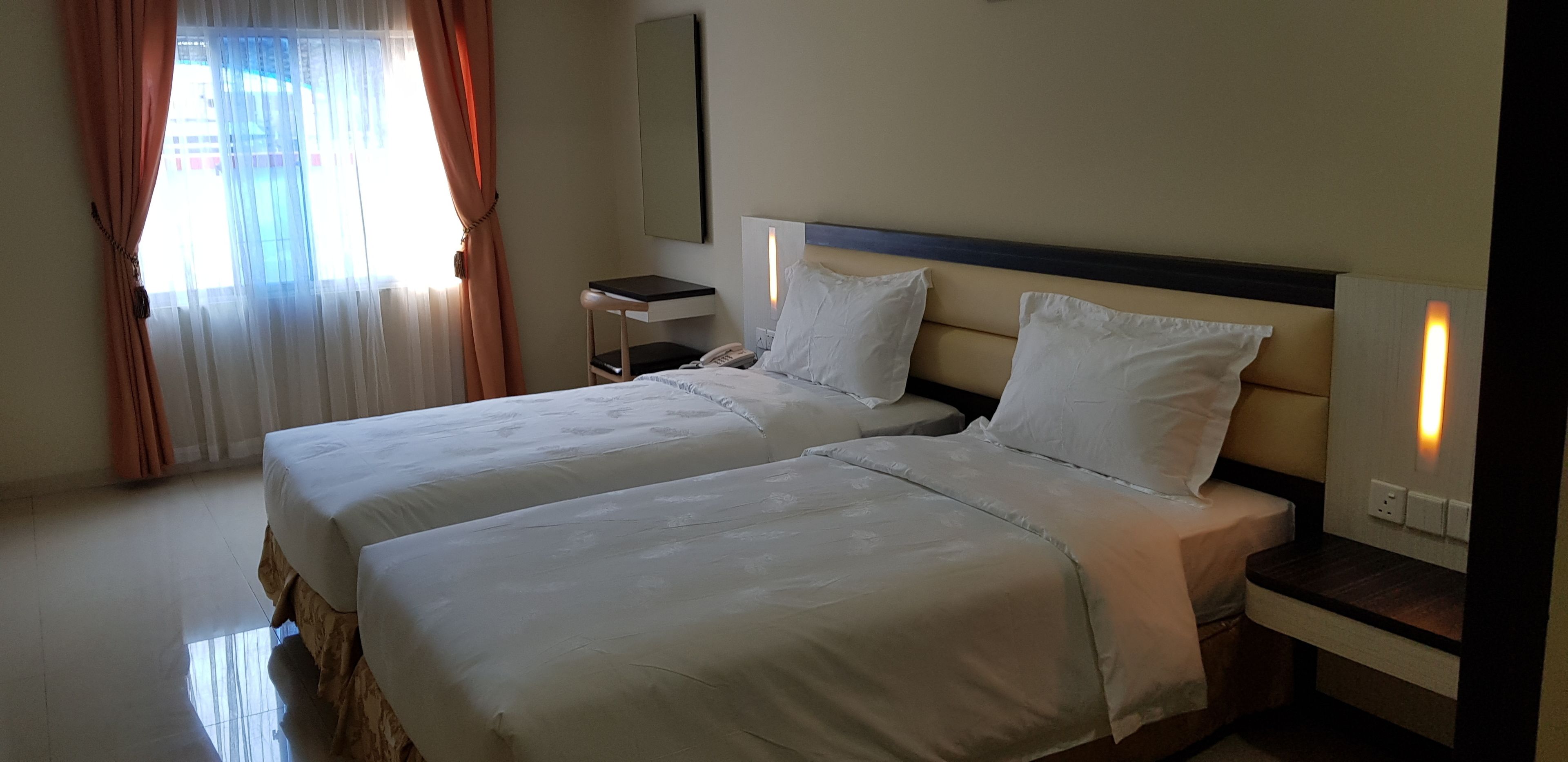 Quarto de hotel com duas camas, uma mesa de cabeceira com telefone, um espelho e uma janela com cortinas.