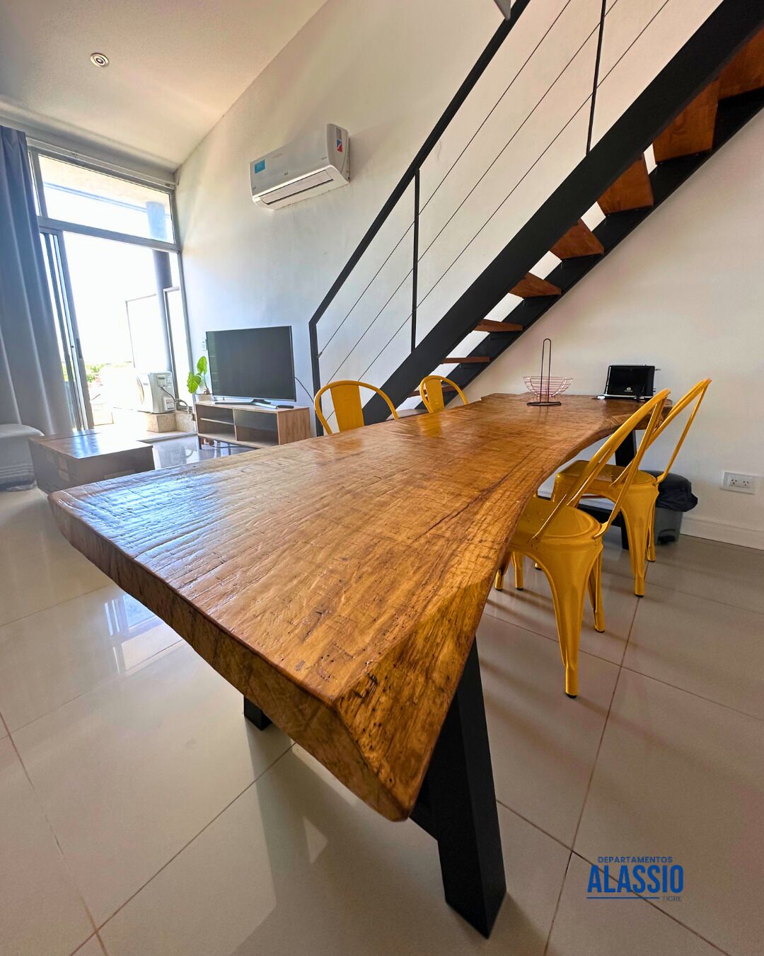 Uma sala de jantar moderna com uma longa mesa de madeira, cadeiras amarelas, uma televisão e uma escada.
