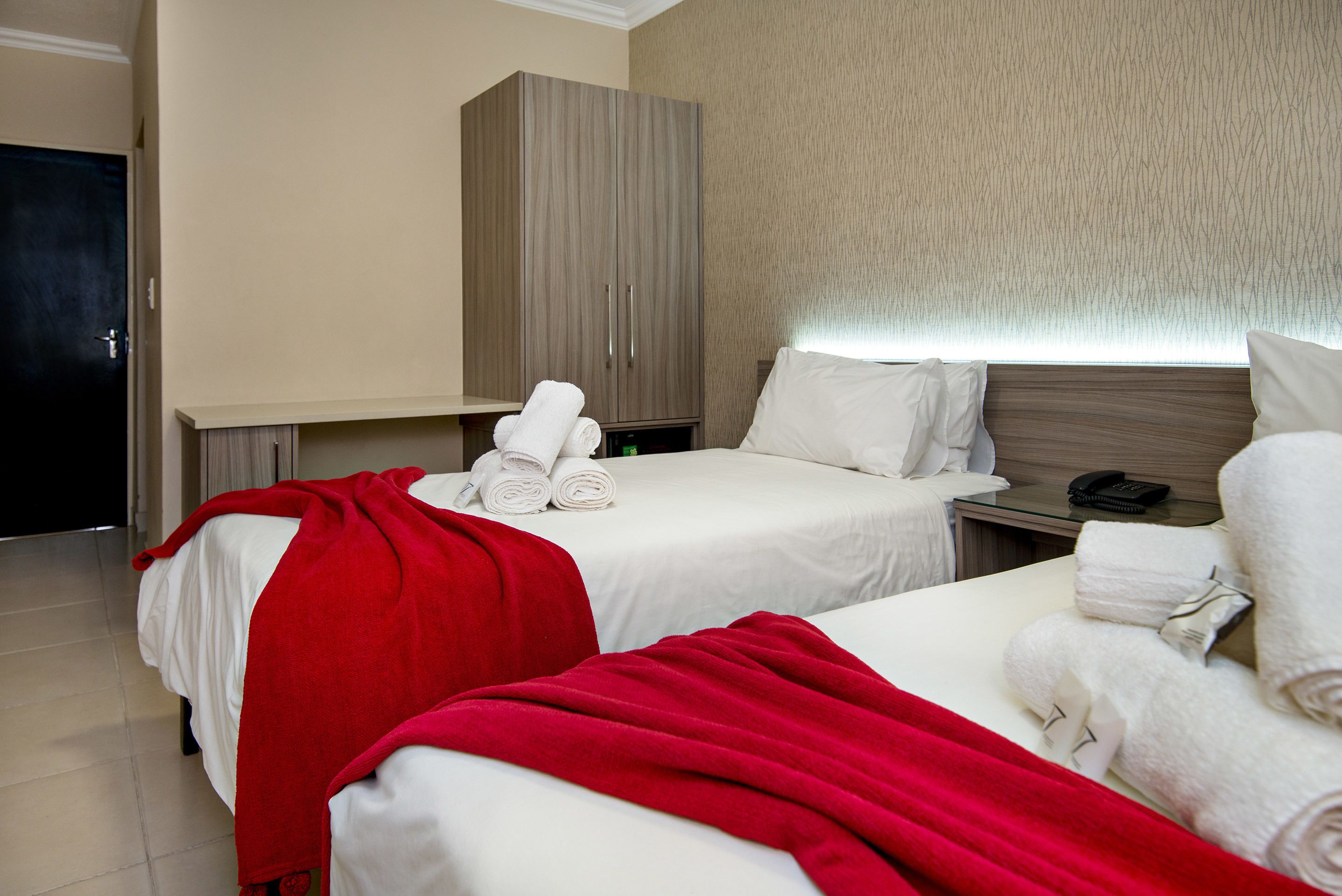 Quarto de hotel com duas camas, cada uma com lençóis brancos e cobertores vermelhos, um guarda-roupa de madeira e uma mesa de cabeceira com um telefone e toalhas.