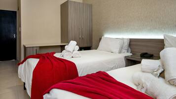 Quarto de hotel com duas camas, cada uma com lençóis brancos e cobertores vermelhos, um guarda-roupa de madeira e uma mesa de cabeceira com um telefone e toalhas.