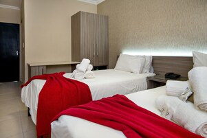 Quarto de hotel com duas camas, cada uma com lençóis brancos e cobertores vermelhos, um guarda-roupa de madeira e uma mesa de cabeceira com um telefone e toalhas.