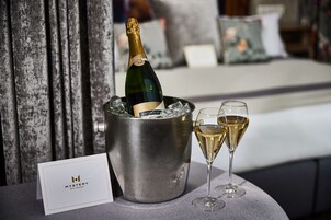 Minibar com uma garrafa de champanhe e duas taças, um cartão branco com o nome do hotel e uma cama com almofadas ao fundo.