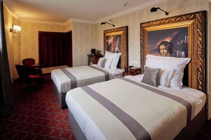 Quarto de hotel com duas camas, uma mesa, uma cadeira e um quadro grande emoldurado na parede.