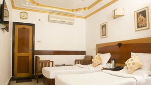 Quarto de hotel com duas camas, cabeceira de madeira, uma mesinha, uma cadeira, um relógio e ar-condicionado fixado na parede.