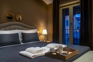 Quarto de hotel com uma cama, duas espelhos, uma mesa de cabeceira com abajur, um suporte com toalhas e chávenas, e vista para varanda.