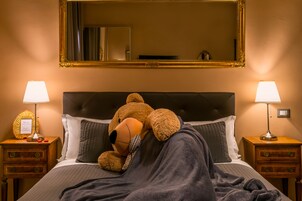 Quarto de hotel com uma cama grande, duas mesas de cabeceira com luminárias, um espelho e um urso de pelúcia na cama.