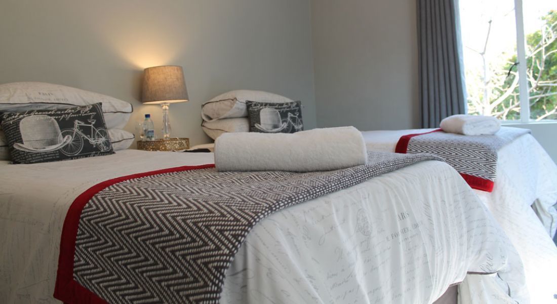 Quarto de hotel com duas camas, cada uma com colcha estampada e almofadas. Há uma mesinha de cabeceira com abajur e uma garrafa de água.