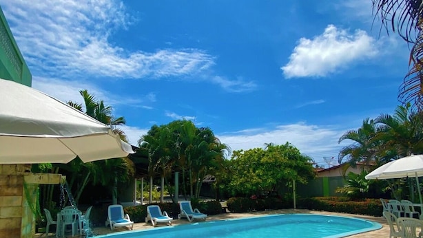 Piscina com espreguiçadeiras e guarda-sóis, cercada por vegetação e céu azul.