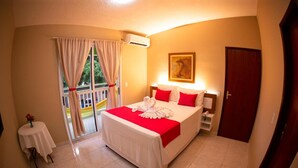 Quarto de hotel com uma cama, uma mesinha com um vaso, uma janela com cortinas e uma porta.