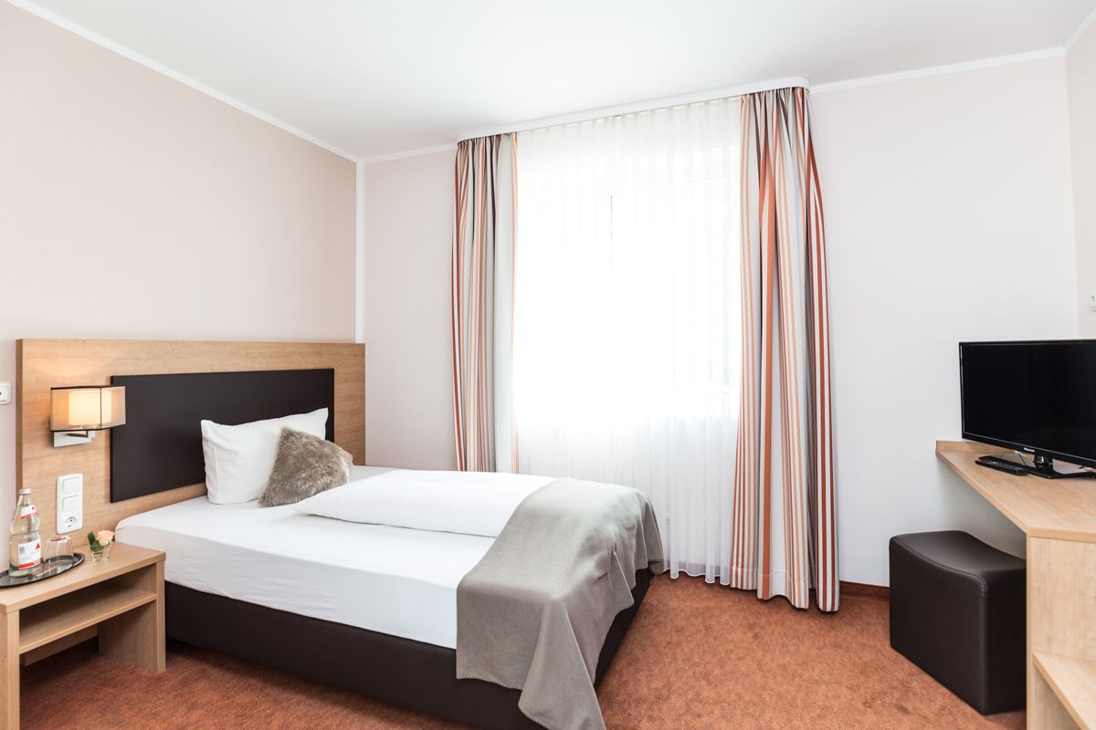 Quarto de hotel com cama, escrivaninha com televisão, mesa de cabeceira e janela com cortinas.