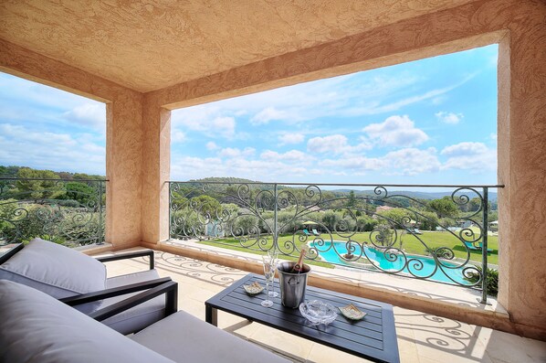 Uma varanda com vista para uma piscina e vegetação, uma mesa com uma taça de vinho e uma garrafa, e um espaço de estar acolchoado.