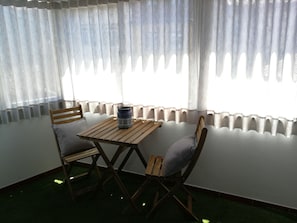 Quarto com uma mesa de madeira e duas cadeiras, um recipiente azul e branco com padrão sobre a mesa, e uma janela com cortinas finas.