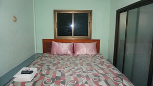 Quarto pequeno com uma cama de solteiro, colcha estampada, cabeceira de madeira, duas almofadas cor-de-rosa, um controle remoto e uma janela com moldura de madeira.