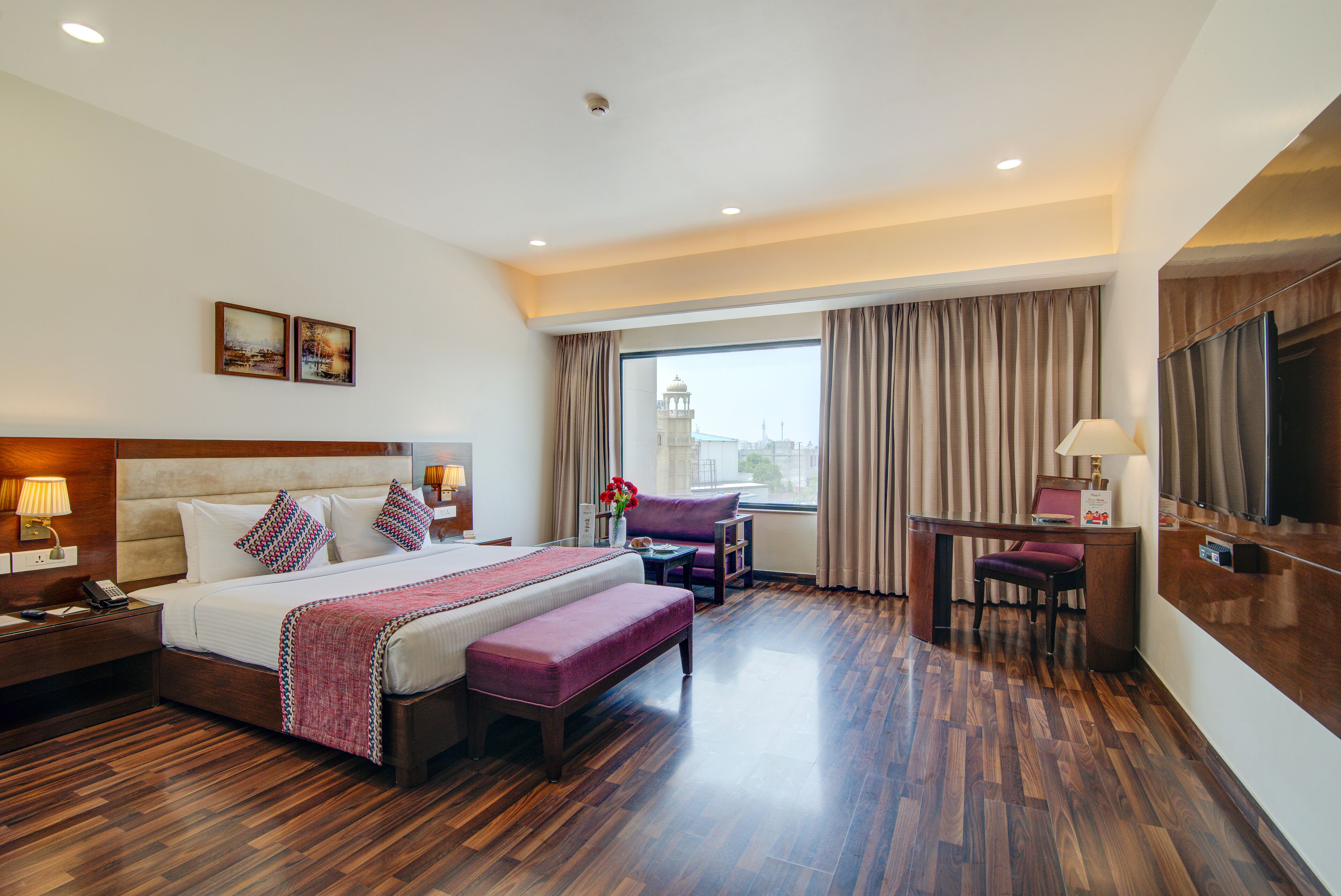 Quarto de hotel com uma cama grande, uma escrivaninha, uma cadeira, uma televisão e uma janela com cortinas.