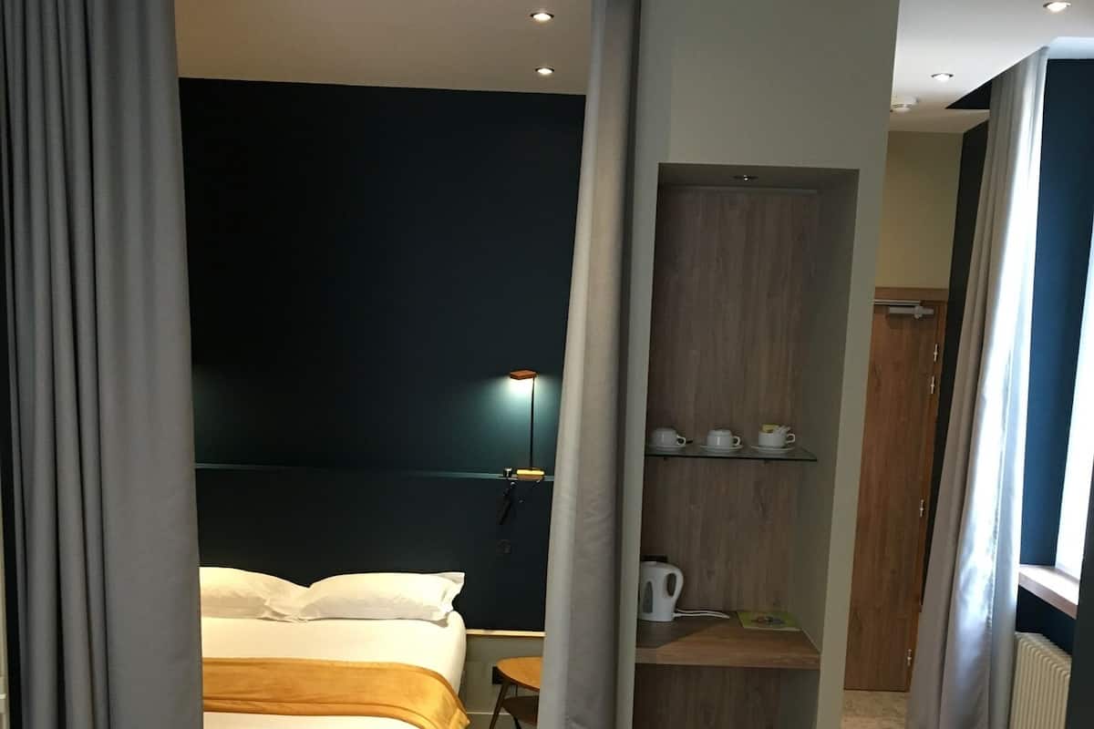 Quarto de hotel com cama, uma mesinha, mesa de cabeceira, prateleira com chávenas e chaleira, e janela com cortinas.