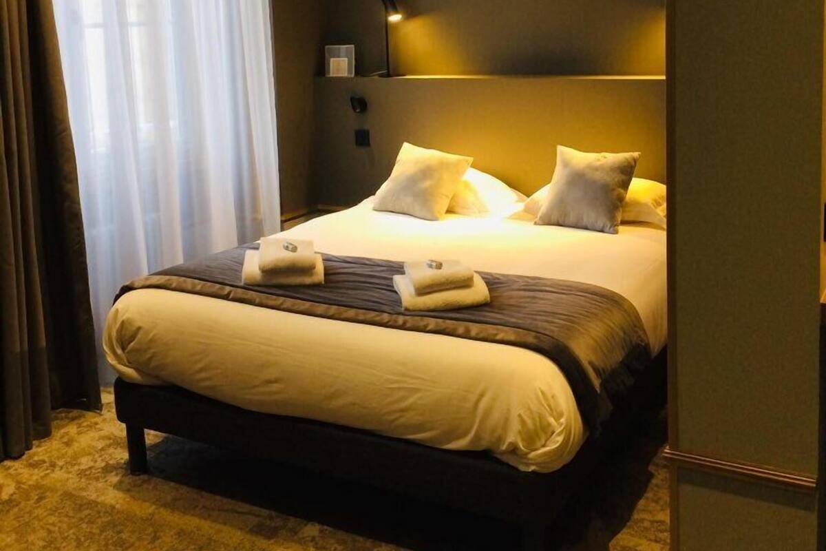 Quarto de hotel com uma cama grande, mesa de cabeceira, abajur e vista para o exterior através de cortinas leves.
