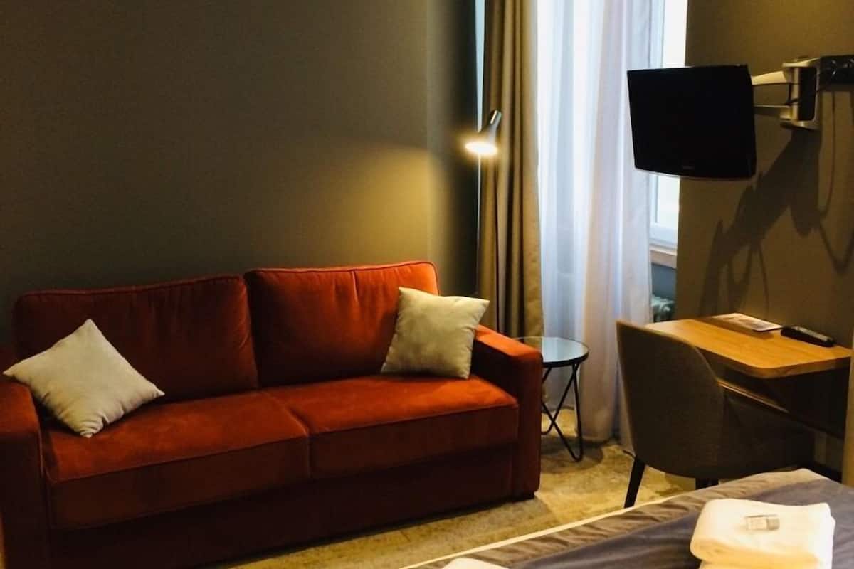 Quarto de hotel com um sofá vermelho, uma escrivaninha com computador, uma televisão na parede e uma cama com toalhas.
