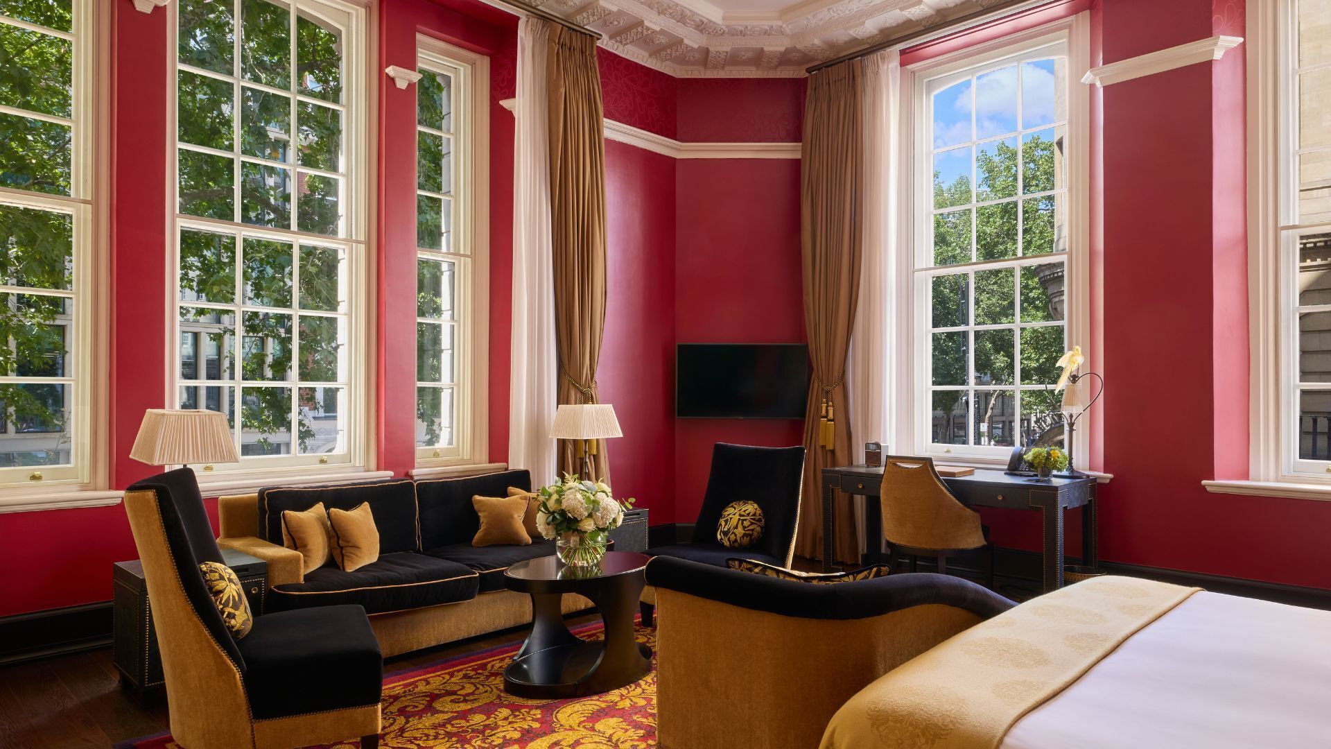 Quarto de hotel com parede vermelha, janelas amplas, sofá, poltrona, cama e televisão.