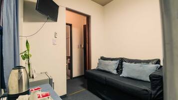 Um quarto pequeno com um sofá de couro preto, uma TV fixada na parede, uma mesinha com chaleira e xícaras, e uma planta.