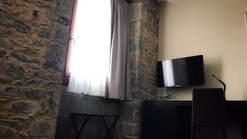 Quarto com parede de pedra, janela com cortinas, televisão de tela plana na parede, uma mesinha e uma cadeira.