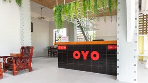 Uma área de recepção com balcão preto, marca 'OYO' em vermelho, plantas penduradas e cadeiras de madeira tradicionais.