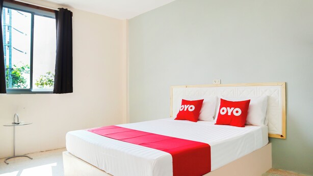 Uma cama bem arrumada com um edredom branco e vermelho, duas almofadas vermelhas com a palavra "OYO" e uma janela com cortinas pretas.