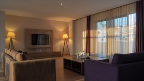 Quarto de hotel moderno com sofá, mesa de centro, TV de tela plana e uma grande janela com cortinas.