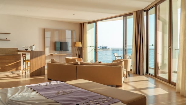 Quarto de hotel moderno com cama grande, sofá, área de jantar e televisão. O quarto oferece vista para o mar e varanda.