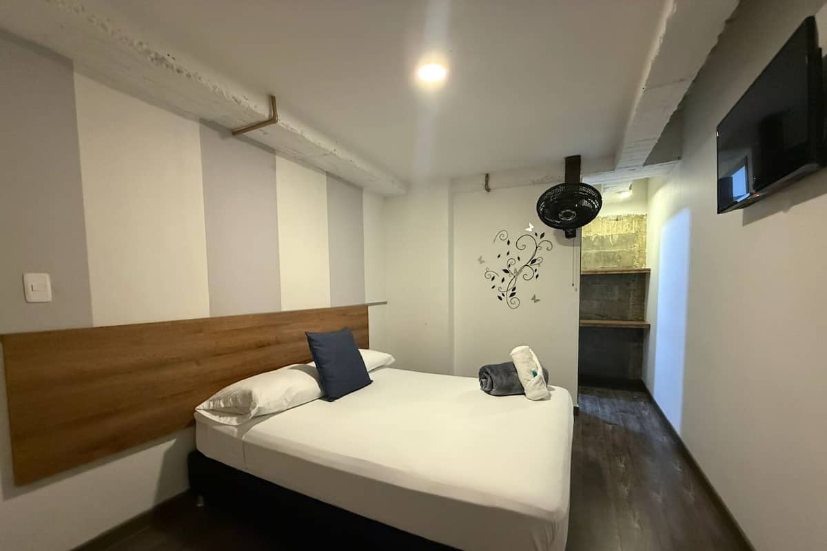 Quarto com cabeceira de madeira, cama com roupa de cama branca, mesinha de cabeceira pequena, televisão fixada na parede e um objeto decorativo na parede.