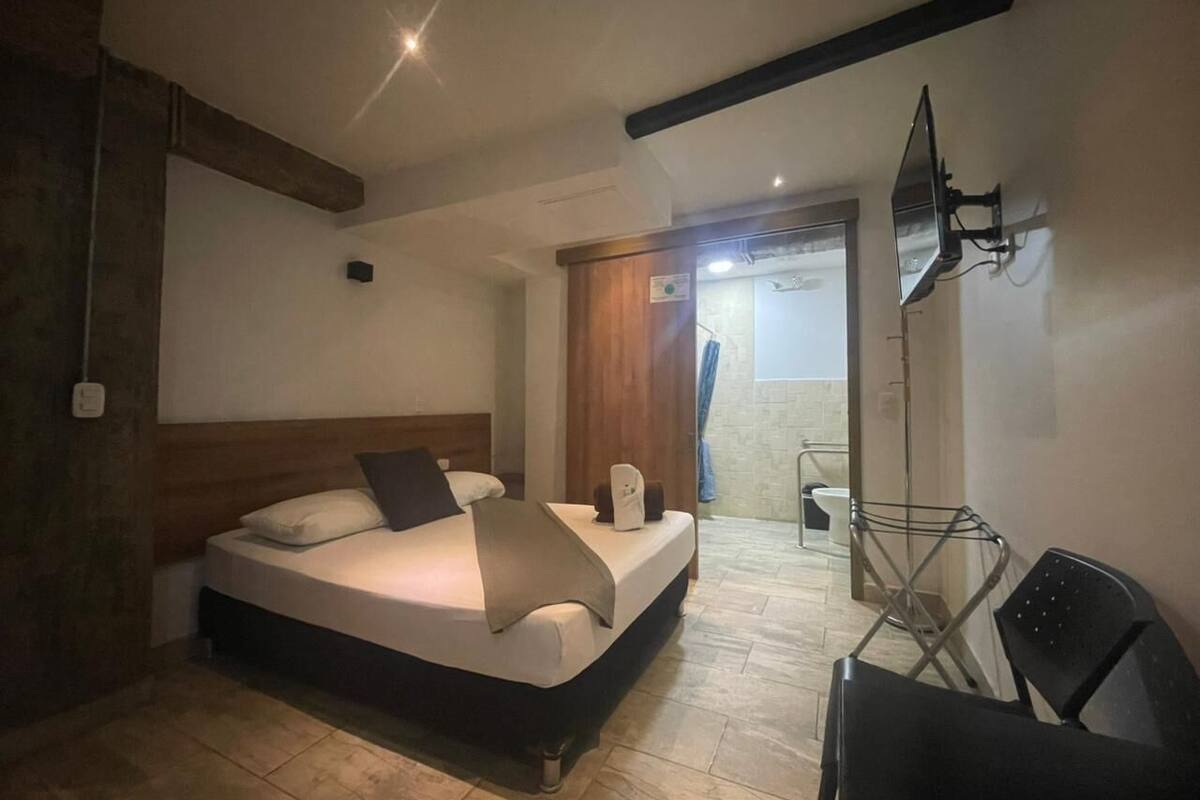 Quarto com uma cama, uma cadeira, uma TV e um banheiro visível ao fundo.