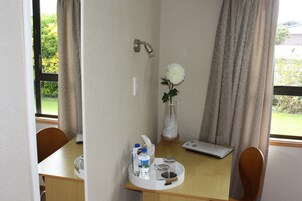 Quarto compacto com chuveiro, uma pequena mesa com artigos de higiene, uma cadeira e uma janela com cortinas.
