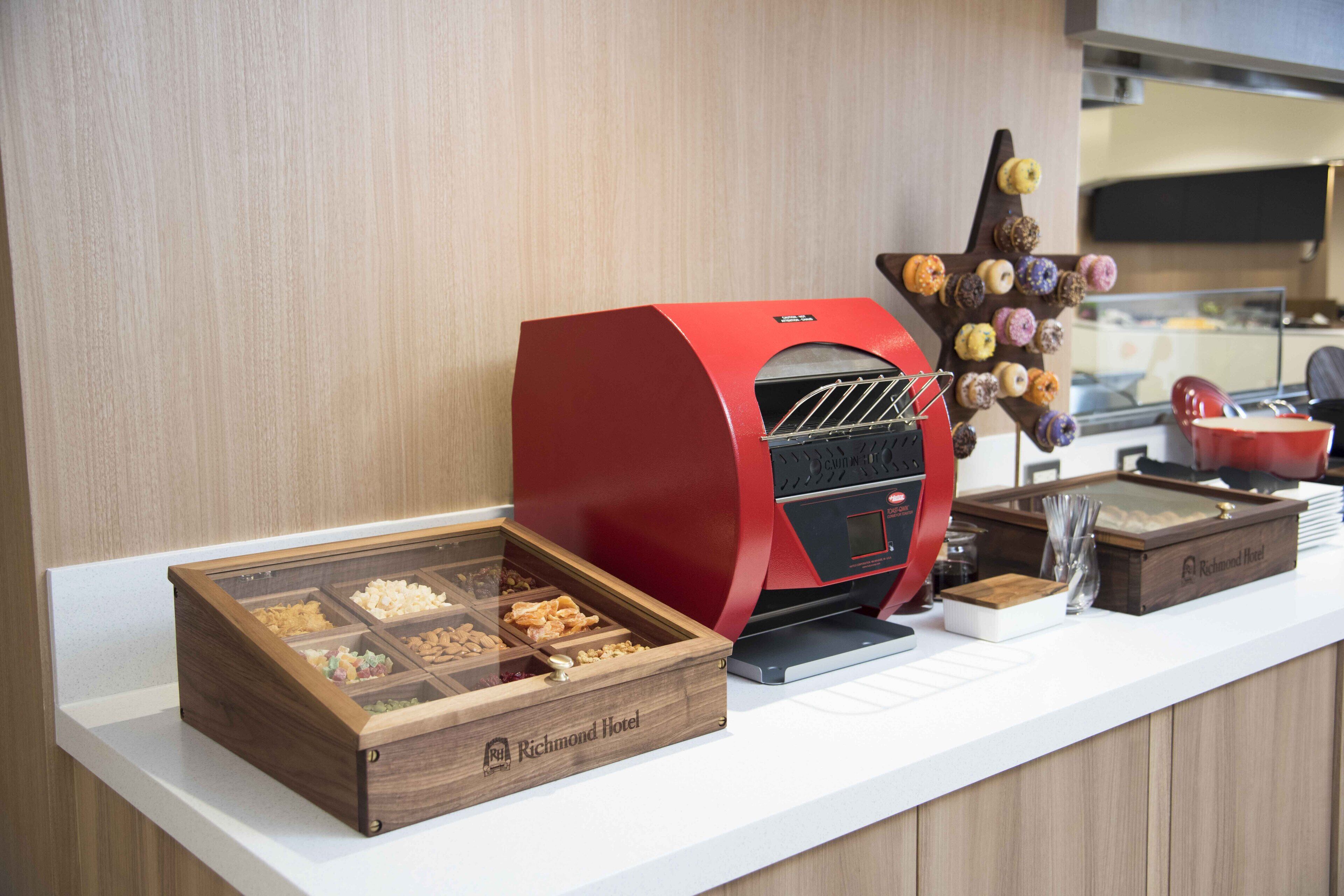 Vitrine de pastelaria com uma máquina de café vermelha, uma caixa de madeira com frutos secos e chocolate, e um expositor de donuts.