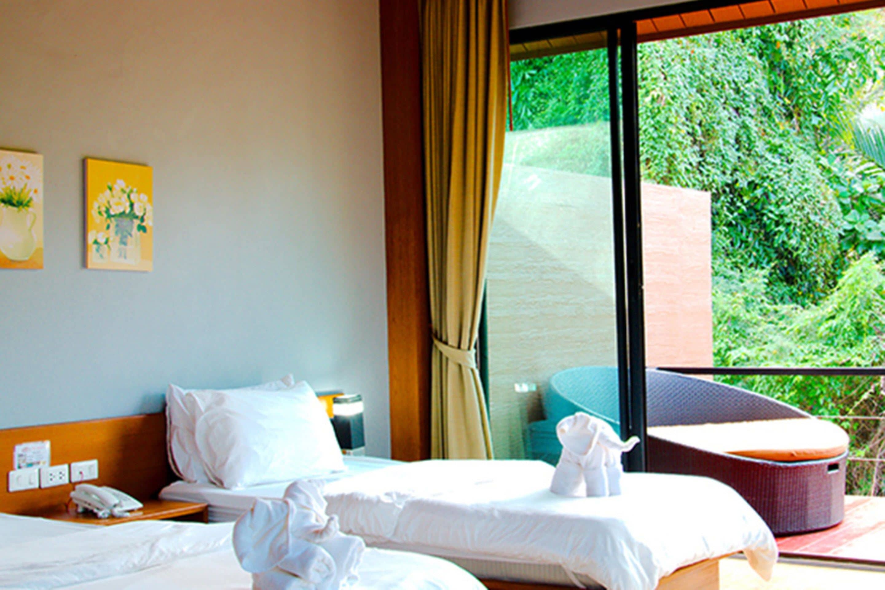 Quarto de hotel com duas camas, uma varanda com cadeira de vime e vista para o verde.