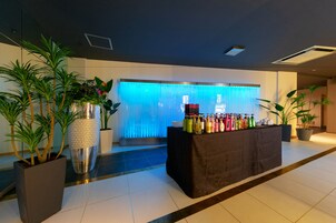 Uma área de recepção com balcão, plantas em vasos, iluminação azul no fundo e diversas bebidas engarrafadas.