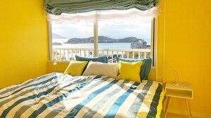 Quarto com cama, almofadas, mesa de cabeceira e janela com vista para a água e terra ao longe.