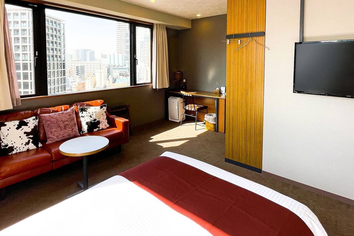 Quarto de hotel com sofá, mesa redonda pequena, cama, televisão, escrivaninha e vista para a cidade pela janela.